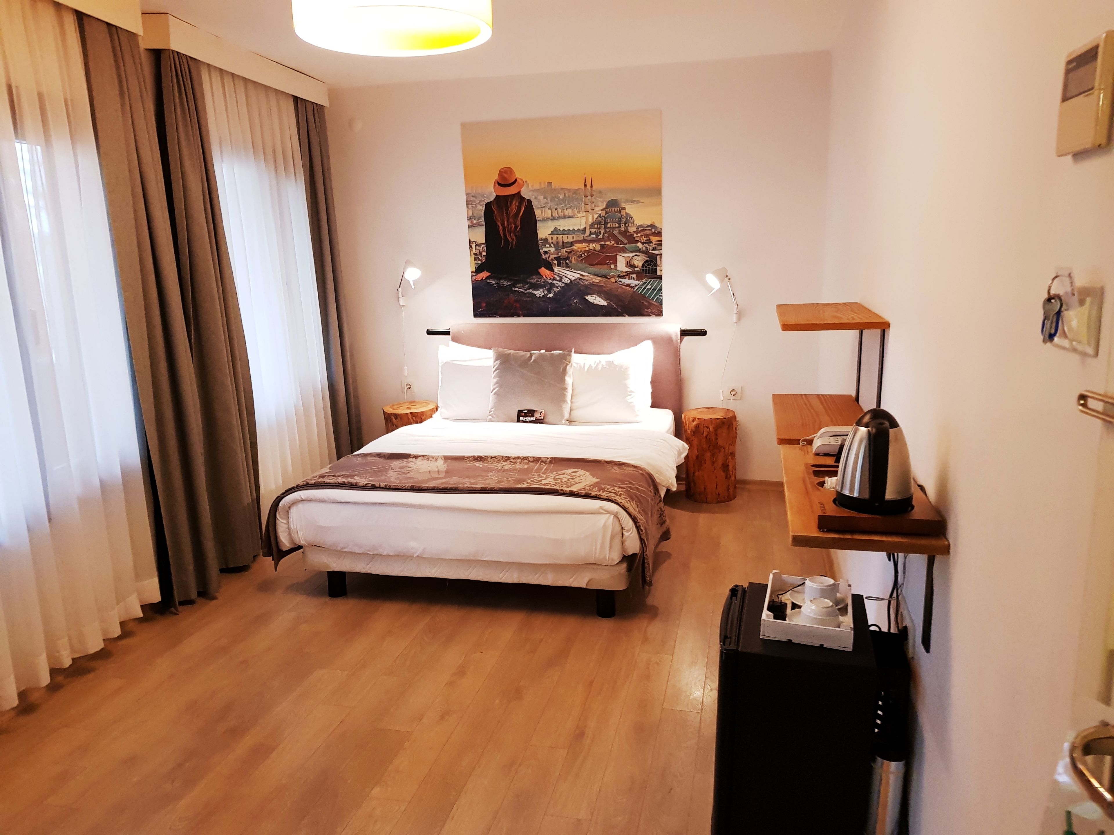 Quarto de hotel com uma cama grande, mesa de cabeceira, mesa lateral de madeira, chaleira e um quadro com vista para a cidade.