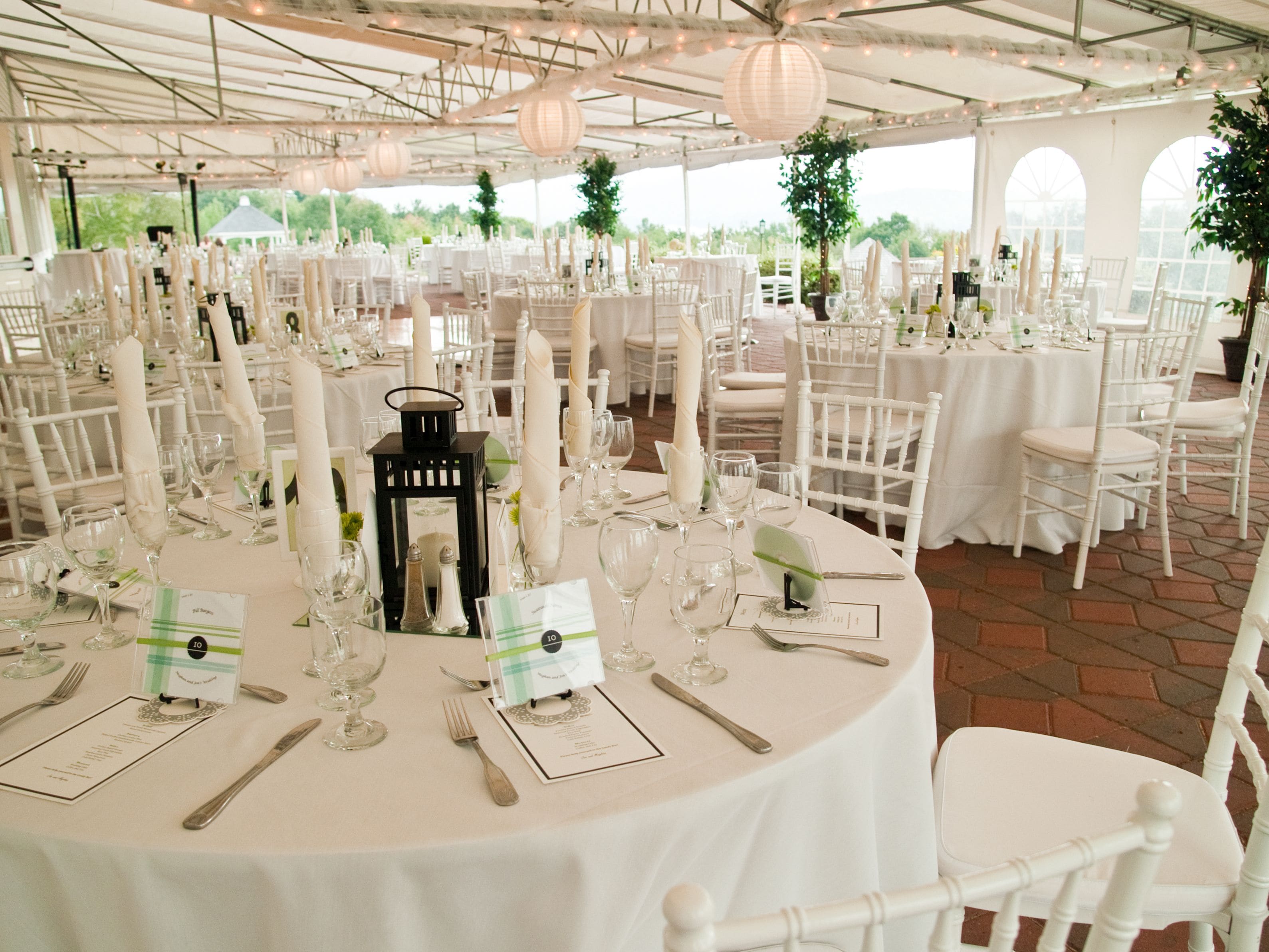 Espaço para eventos ao ar livre com tendas, mesas redondas preparadas para refeições, cadeiras brancas e centros de mesa com velas.