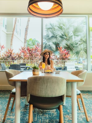 Espaço de refeições com mesa, cadeiras, planta em vaso e vista para plantas tropicais lá fora.