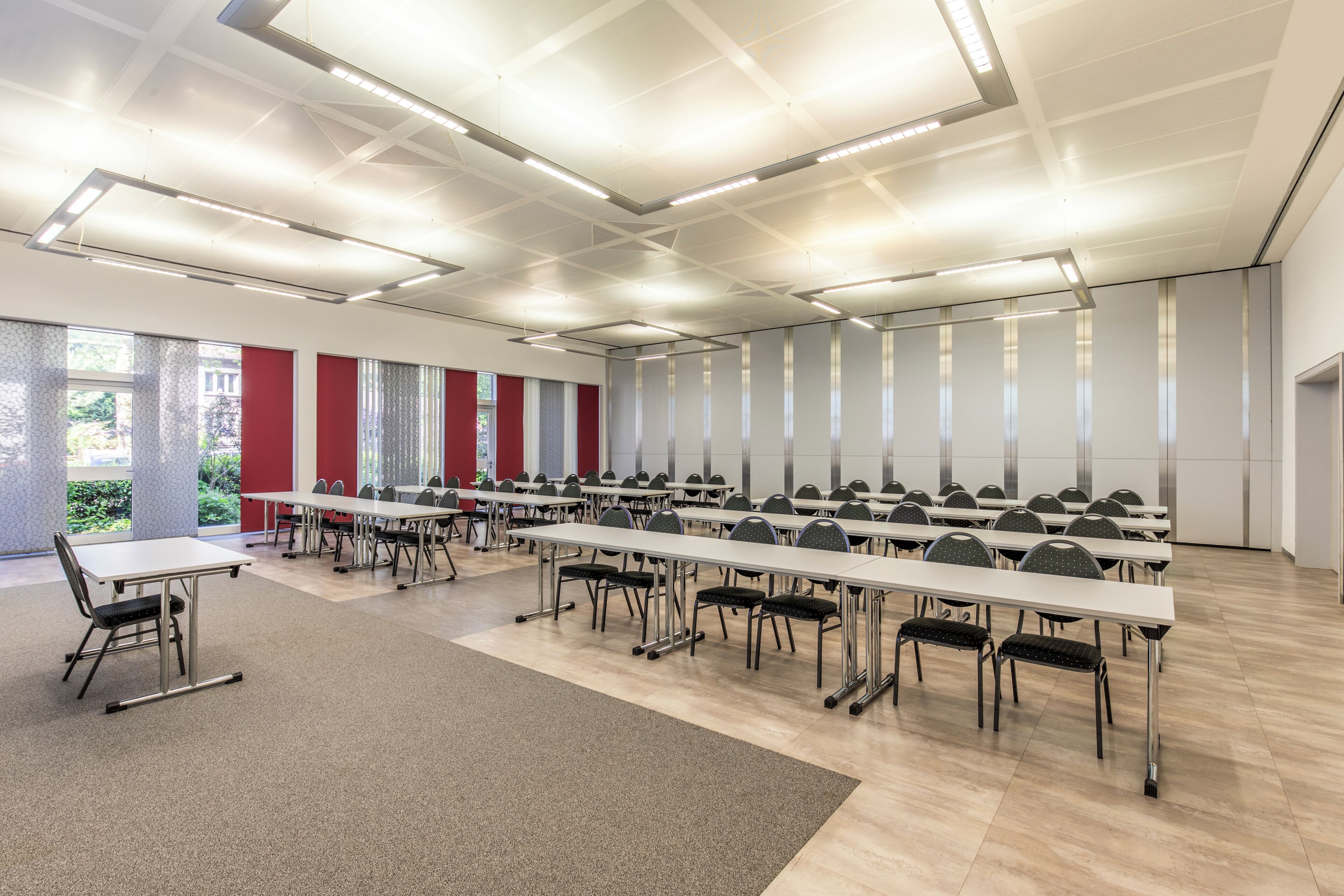 Uma sala de conferências com várias mesas e cadeiras, janelas amplas e teto alto com luminárias.