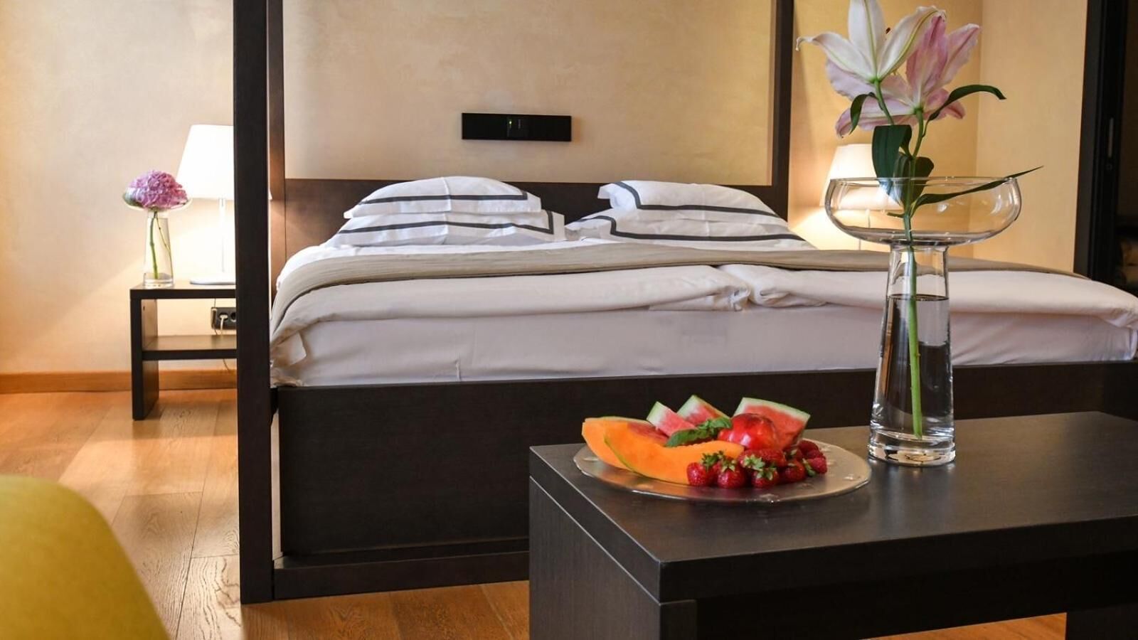 Quarto de hotel com cama bem arrumada, mesa de cabeceira com um prato de frutas e um vaso com flores.