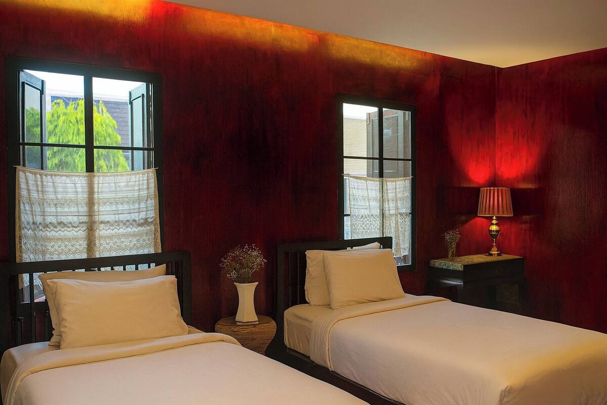 Quarto com duas camas, janela com cortinas de renda, mesa de cabeceira com abajur e vista para o verde lá fora.