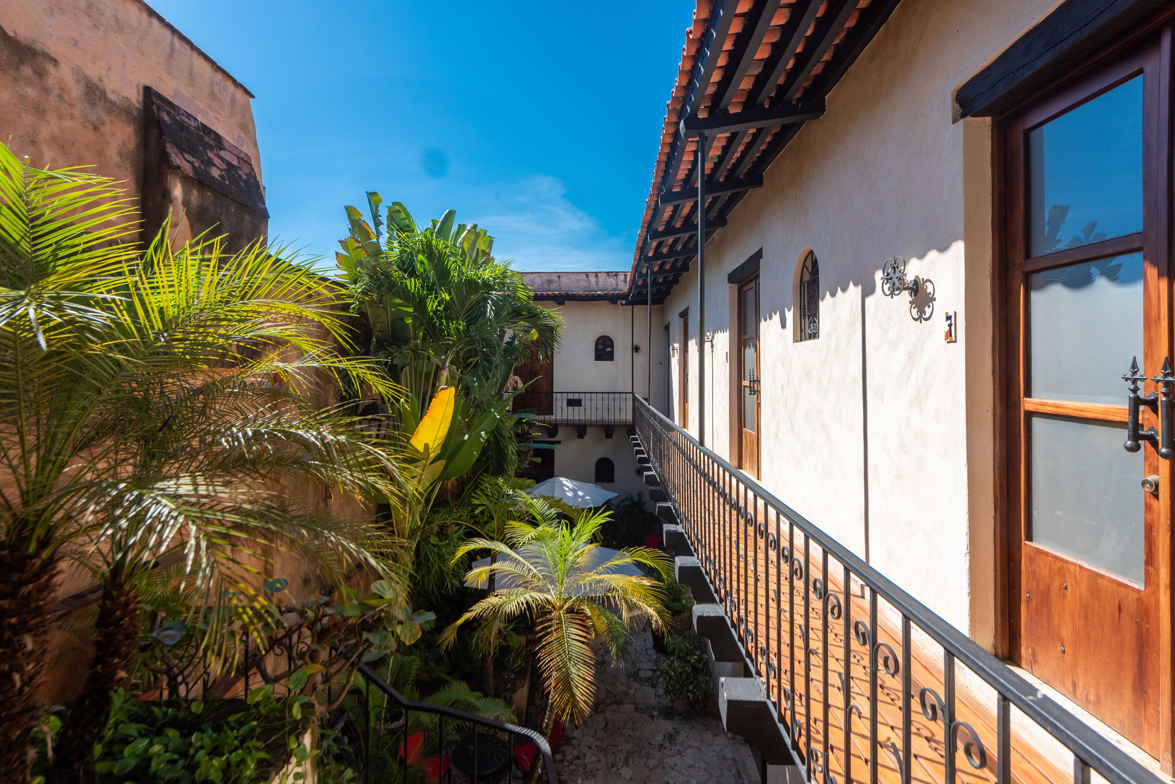 Um pátio estreito com uma escadaria de madeira, um edifício branco com varanda e vegetação exuberante.