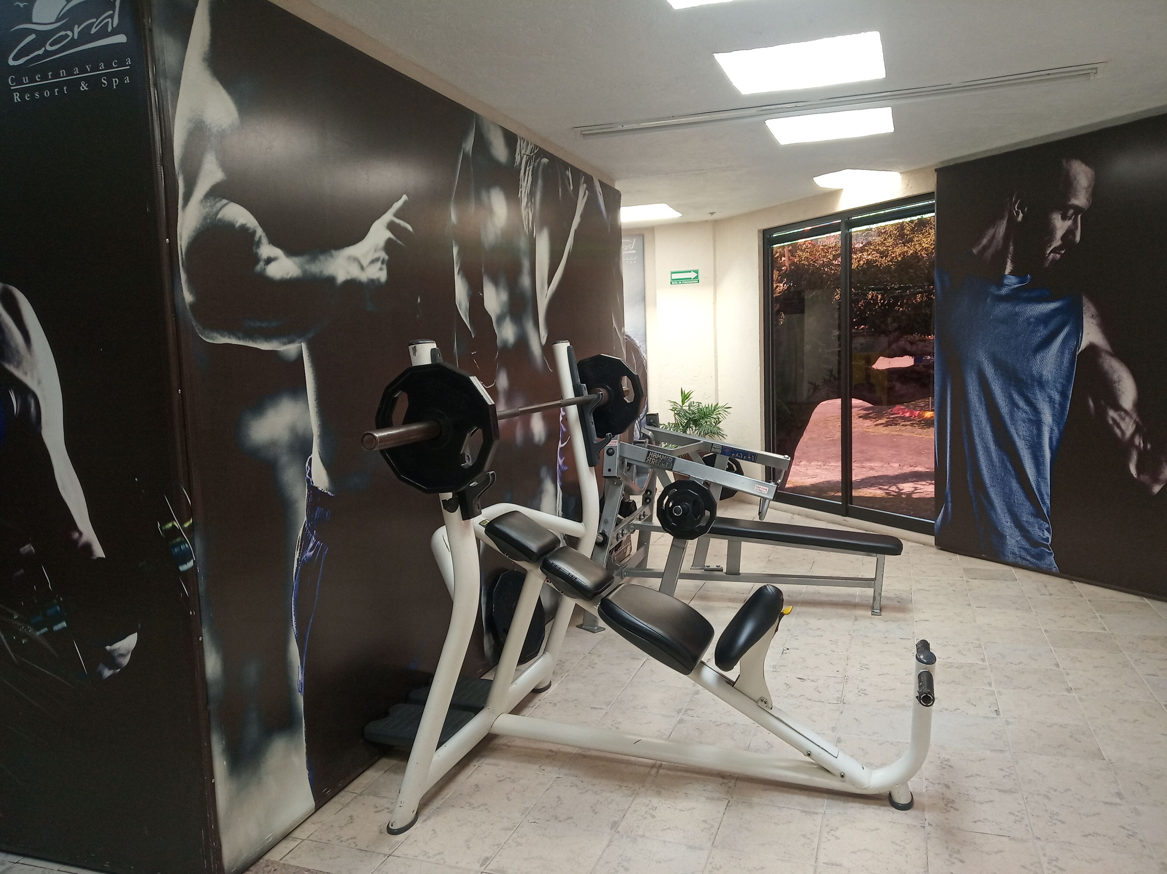 Uma academia com equipamentos de musculação, incluindo uma barra de supino, e uma parede decorada com cartazes sobre fitness.