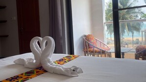 Quarto de hotel com cama, uma janela grande com vista para palmeiras e uma praia, e uma cadeira com almofadas coloridas.