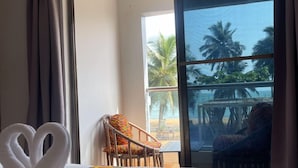 Quarto de hotel com uma cama, janela com vista para palmeiras e praia, e uma cadeira junto à janela.