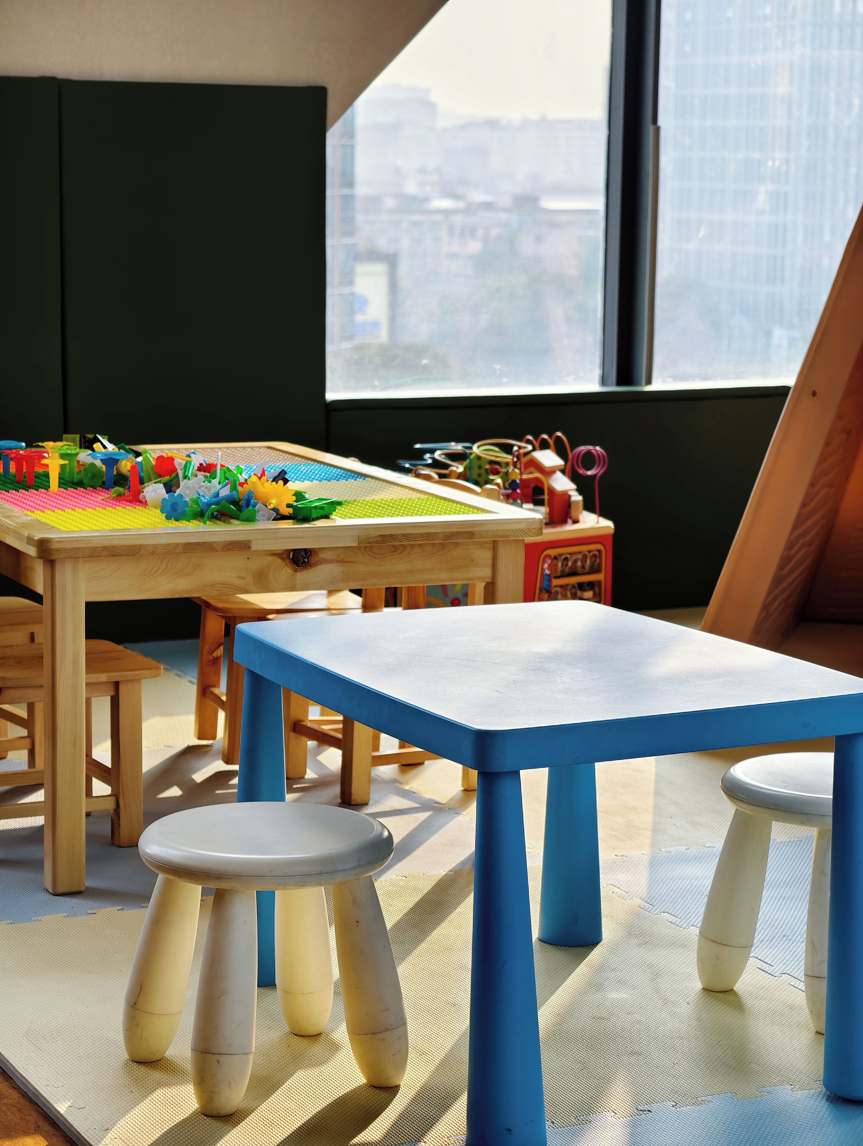 Um quarto com uma janela grande, uma mesa de madeira com brinquedos coloridos e uma mesa azul.