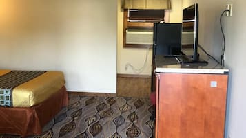 Quarto de hotel com cama, escrivaninha com computador, lixeira e janela com persianas.