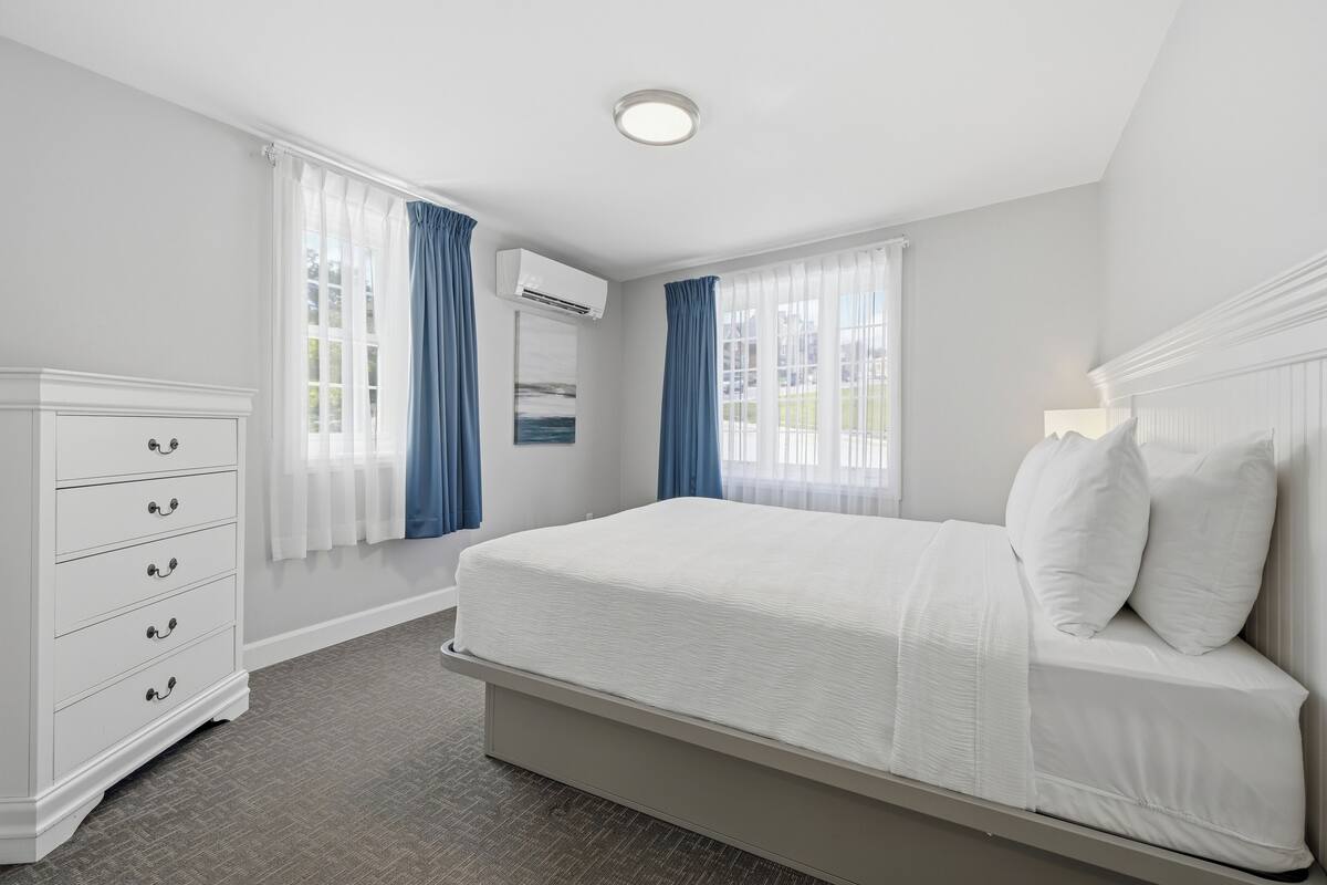 Um quarto com cama, cômoda, ar condicionado e janela com cortinas.