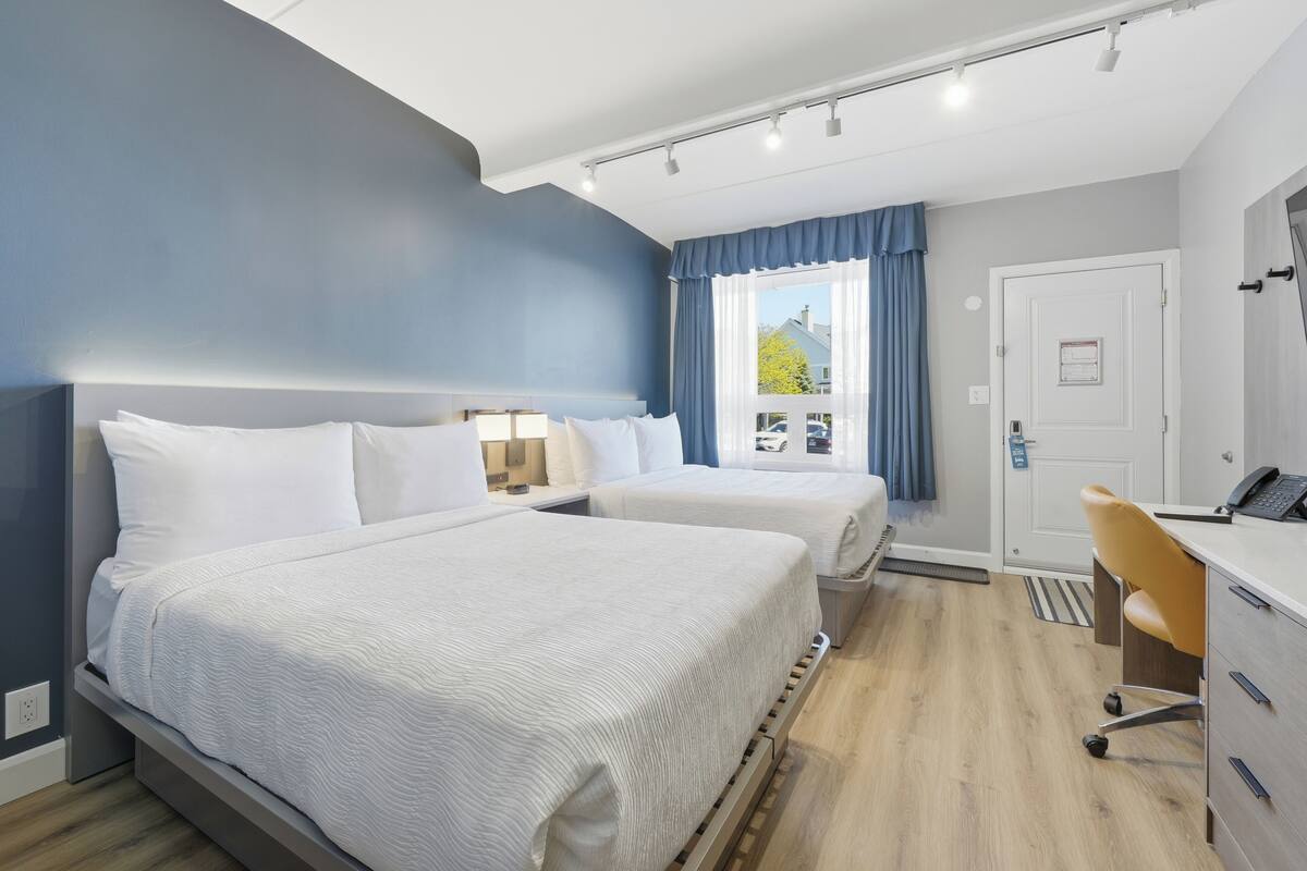 Quarto de hotel com duas camas, uma mesa, uma cadeira, uma janela com cortinas e uma porta com placa.