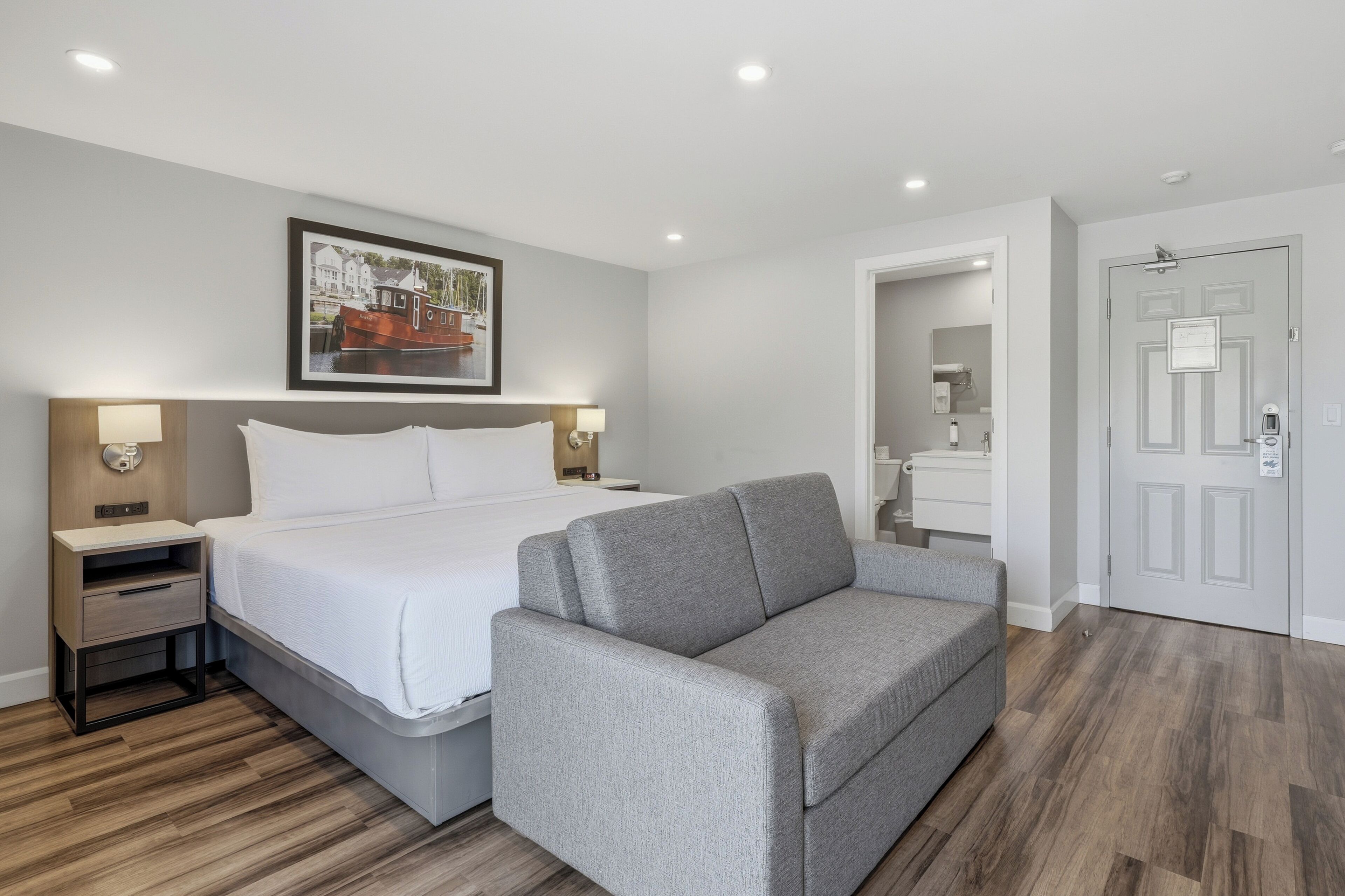 Quarto de hotel com cama, sofá cinza, mesa de cabeceira, quadro na parede e uma porta.