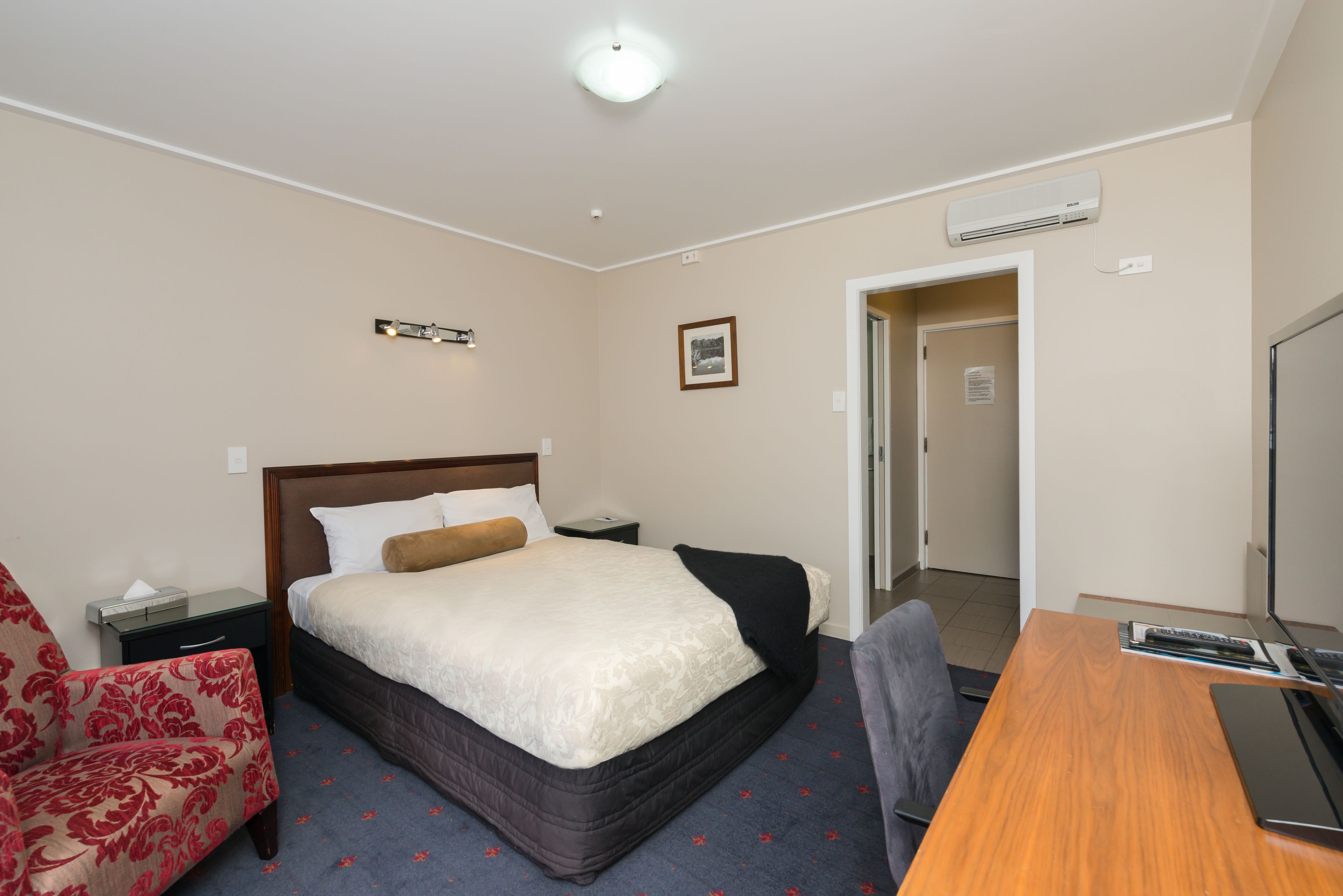 Quarto de hotel com cama, escrivaninha, cadeira, mesa de cabeceira e ar condicionado fixado na parede.