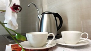 Uma cozinha com chaleira de aço inoxidável, duas xícaras brancas em pires e uma única orquídea branca.