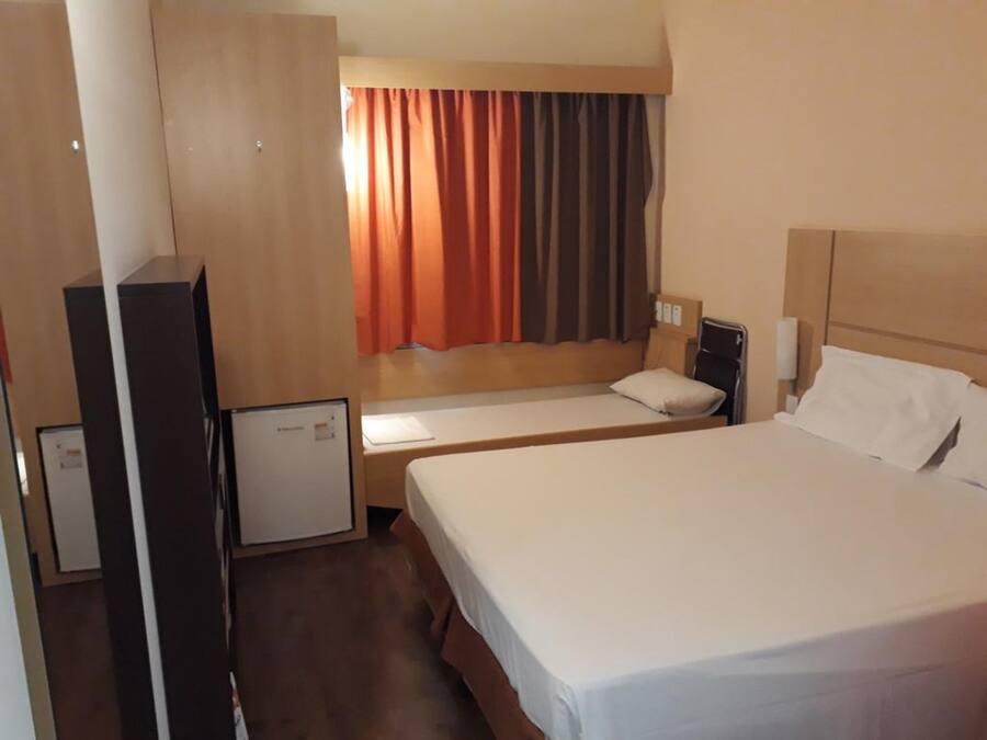 Quarto de hotel com uma cama de solteiro, escrivaninha, cadeira, mini-geladeira e janela com cortinas.