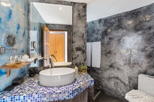 Uma casa de banho moderna com uma grande pia oval, uma prateleira de madeira com artigos de higiene pessoal, uma parede de mármore e uma sanita branca.