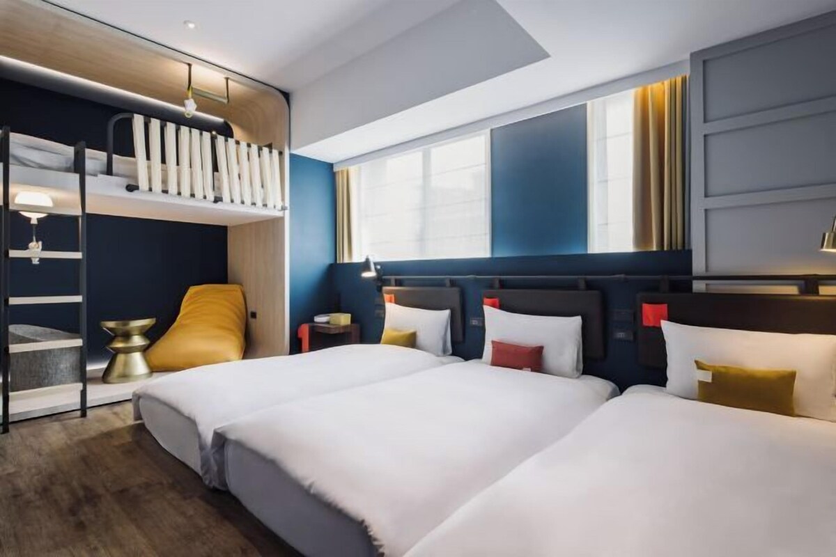 Quarto de hotel moderno com duas camas, um banco embutido, uma janela com cortinas e uma mesa de cabeceira com abajur.