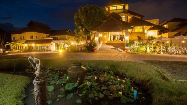 Entrada de hotel bem iluminada, com lago, degraus que levam ao edifício principal e uma área coberta com guarda-sóis.