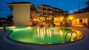 Piscina com tonalidade esverdeada, rodeada por espreguiçadeiras e guarda-sóis, com edifícios ao fundo.