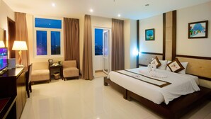 Quarto de hotel com uma cama grande, uma escrivaninha com computador, uma cadeira e vista para a cidade à noite.
