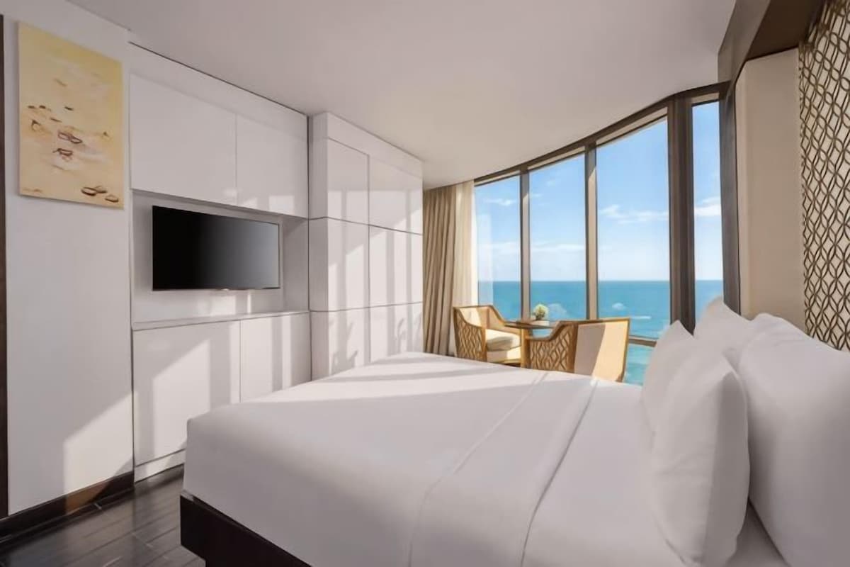 Quarto de hotel com uma cama grande, televisão de tela plana, área de jantar com mesa e cadeiras, e vista para o oceano.