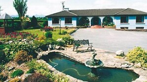 Um edifício de dois andares com fachada azul e branca, um pequeno lago com uma fonte e um jardim com diversas plantas e flores.