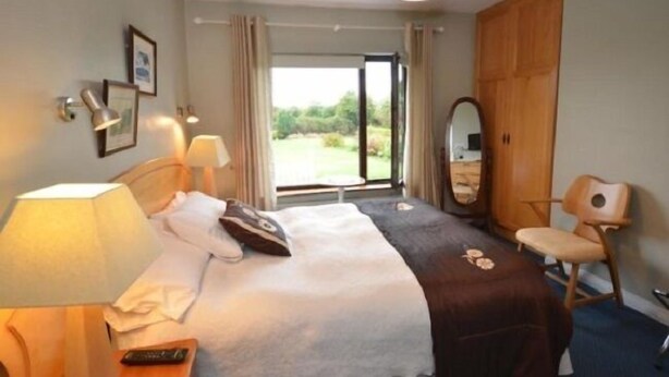 Quarto de hotel com cama, escrivaninha com luminária, cadeira, guarda-roupa e uma grande janela com vista para área verde.