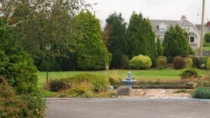 Um jardim residencial com uma fonte, sebes bem aparadas e uma casa ao fundo.