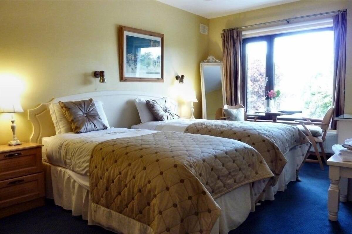 Quarto de hotel com duas camas, uma escrivaninha, uma cadeira, um abajur e uma janela com cortinas.