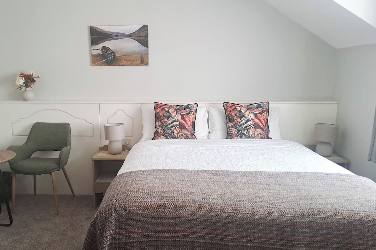 Quarto de hotel com uma cama, uma cadeira, uma mesinha, uma mesa de cabeceira, um abajur e um quadro na parede.