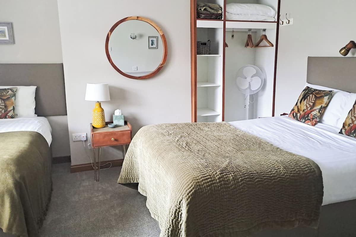 Quarto de hotel com duas camas, uma mesa de cabeceira com luminária, um espelho, um guarda-roupa e um ventilador.