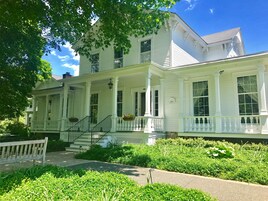 Uma casa branca de dois andares com varanda frontal, escadas e um jardim da frente com vegetação.