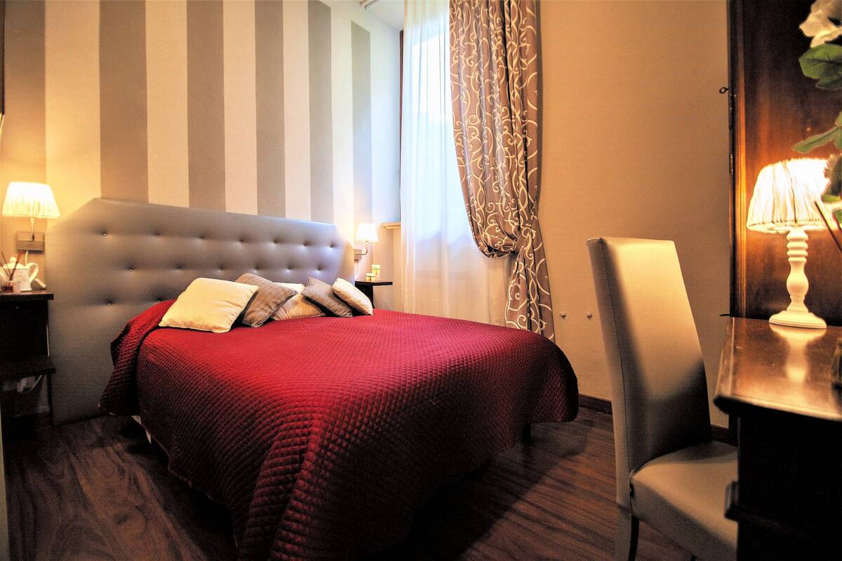 Quarto de hotel com uma cama grande, uma cadeira, uma mesinha de cabeceira e um abajur.