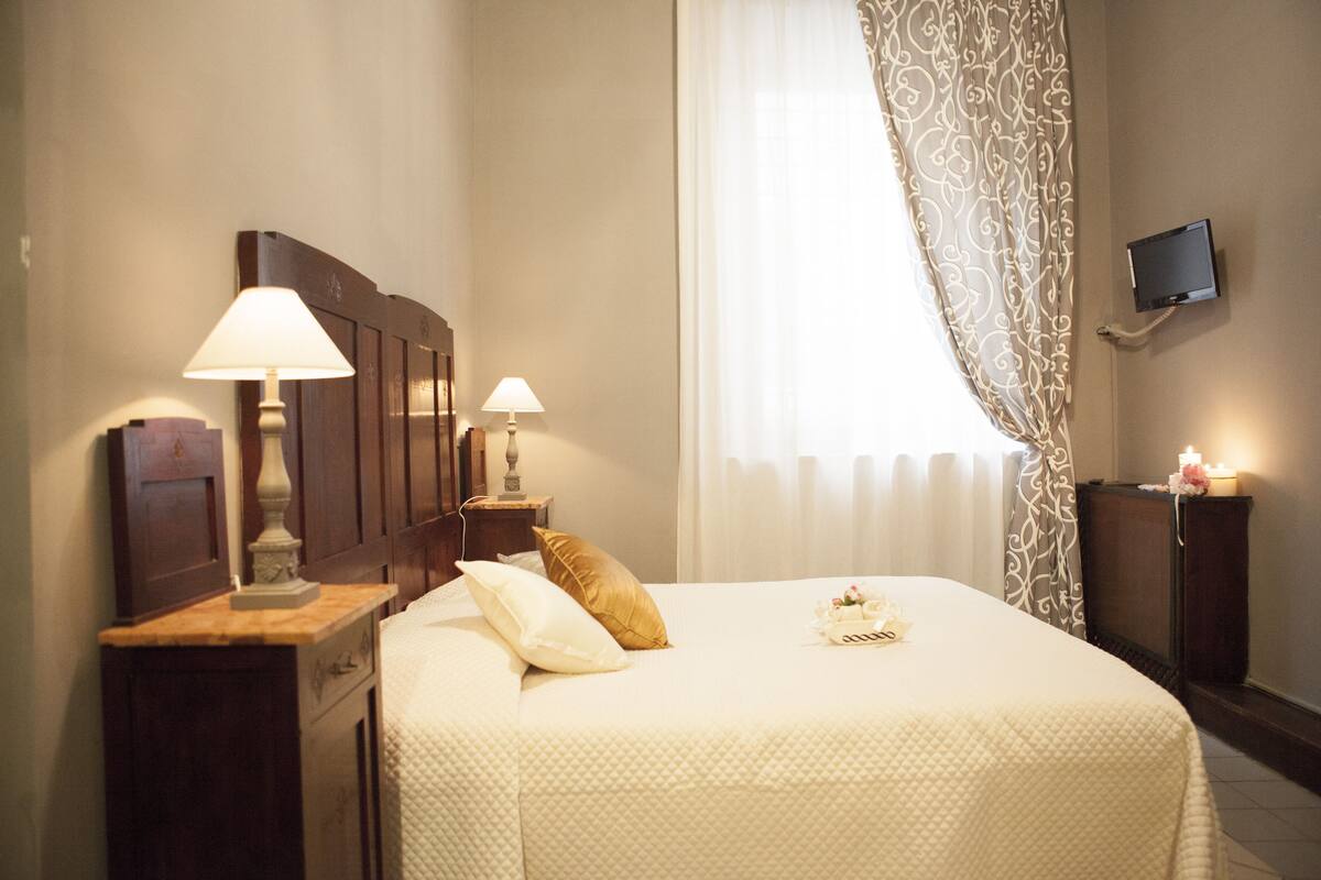 Uma cama bem arrumada com colcha branca, duas luminárias de cabeceira, um criado-mudo de madeira, uma televisão montada na parede e uma janela com cortina estampada.