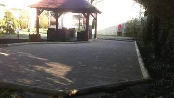Um gazebo com telhado vermelho, rodeado por um espaço pavimentado e uma faixa gramada.