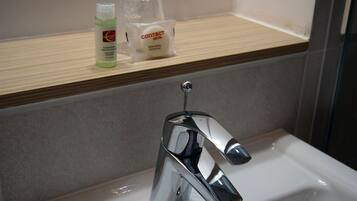 Uma pia de banheiro com torneira cromada, uma garrafa de shampoo e um sabonete de hotel em barra sobre uma prateleira de madeira.
