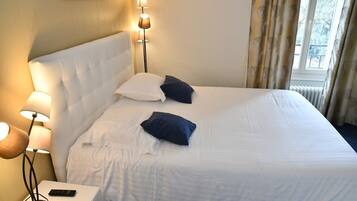 Uma cama bem arrumada com almofadas azuis e brancas, uma luminária de cabeceira e um controle remoto sobre um criado-mudo branco.