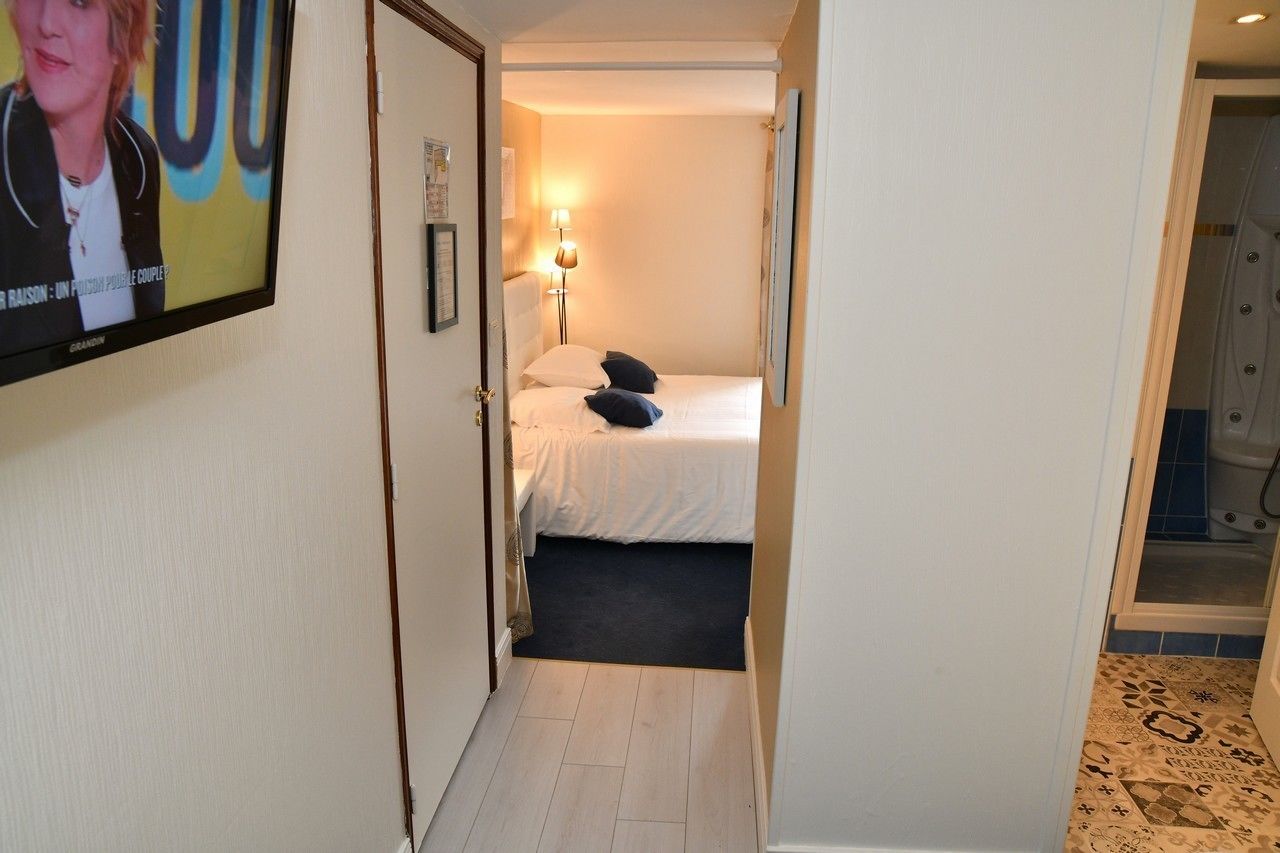 Corredor estreito que dá para um quarto com cama, mesa de cabeceira, abajur e televisão na parede.
