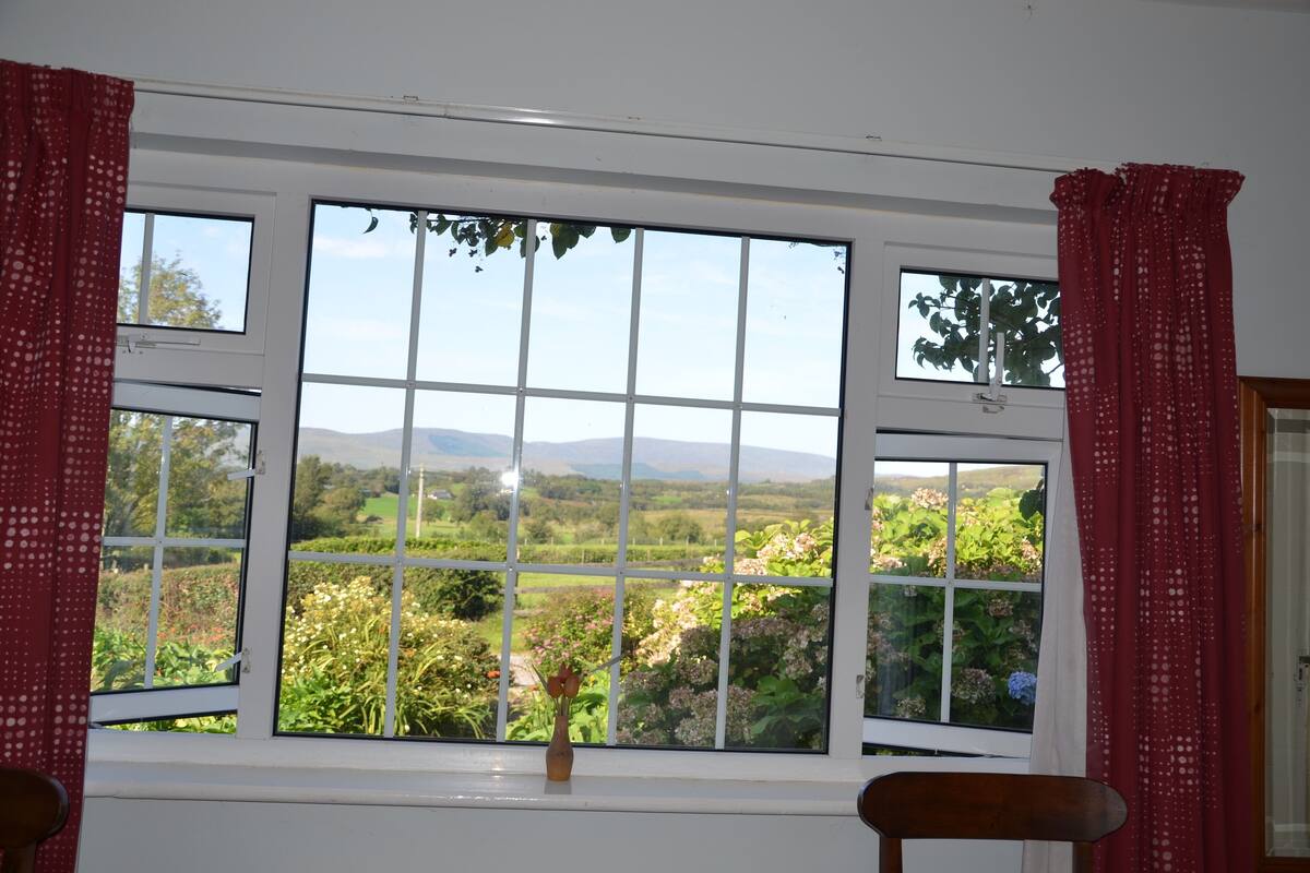 Quarto com uma grande janela que oferece vista para um jardim e montanhas.