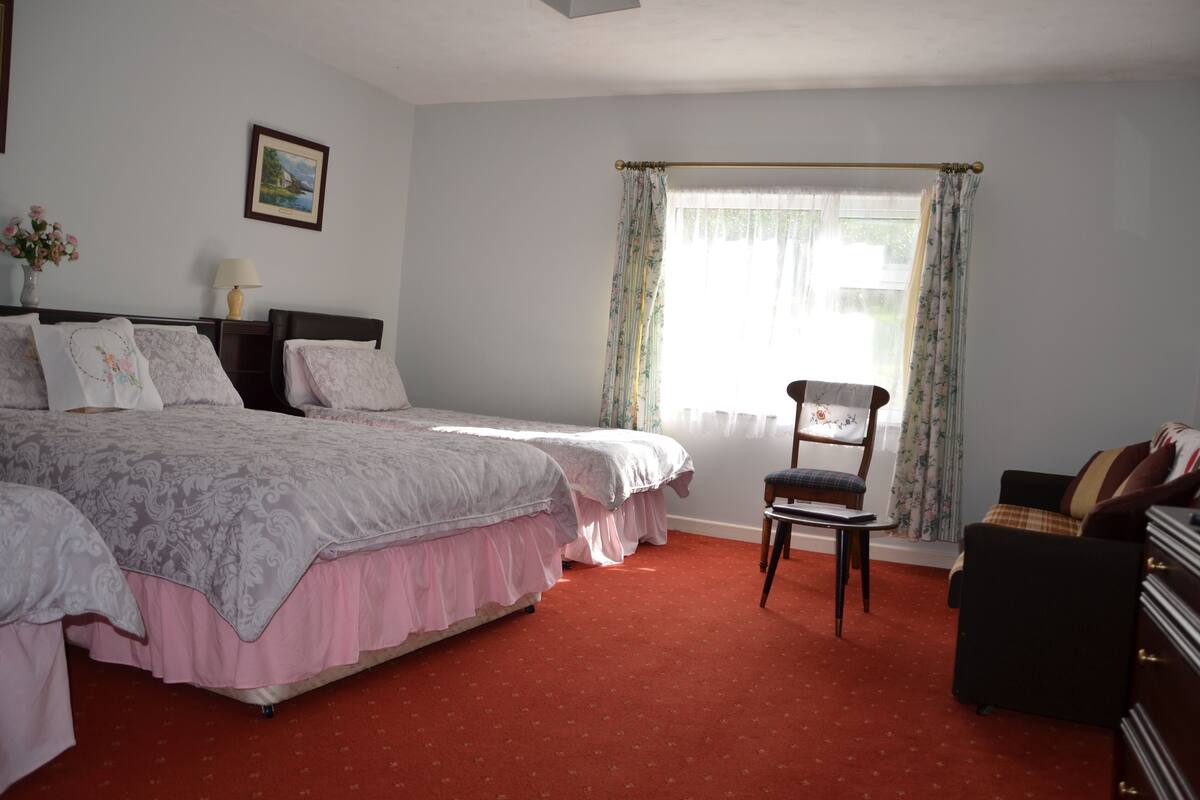 Quarto de hotel com duas camas, uma cadeira, cômoda, abajur e janela com cortinas.