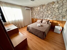 Quarto com parede de pedra, piso de madeira, cama com colcha marrom, mesa de cabeceira com abajur e um aparelho branco pequeno.