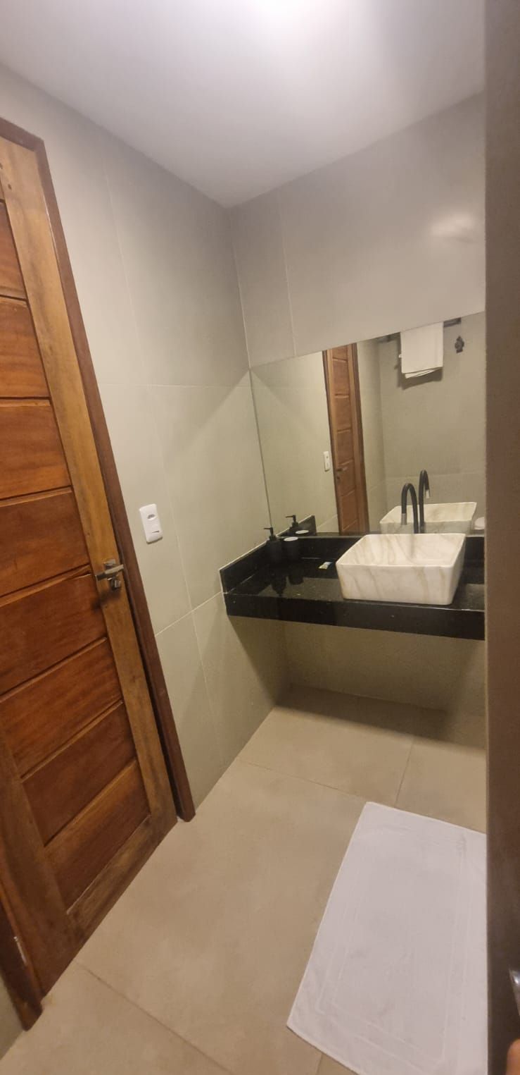 Um banheiro com porta de madeira, bancada preta, pia branca e espelho.
