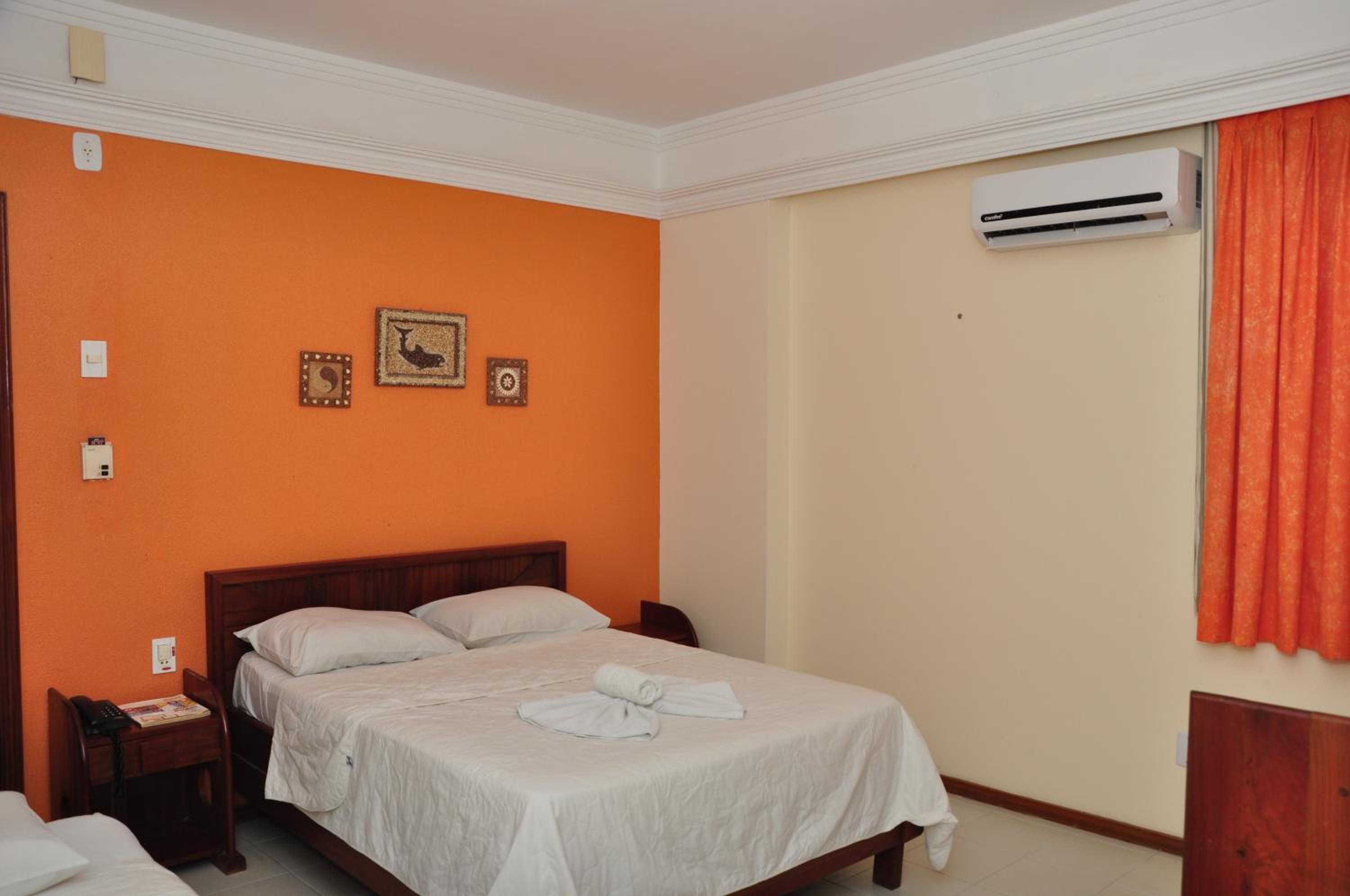 Quarto de hotel com parede laranja, cama de madeira, roupa de cama branca, mesa de cabeceira com telefone e unidade de ar condicionado.