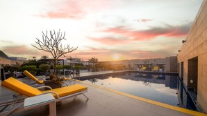 Piscina ao ar livre com cadeiras de descanso amarelas, uma árvore e um pôr do sol ao fundo.