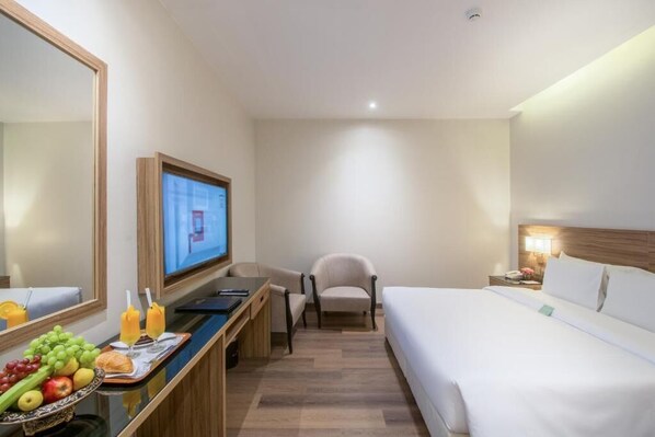 Quarto de hotel com cama, escrivaninha com computador, cadeira, televisão e bandeja de café da manhã com frutas e suco.