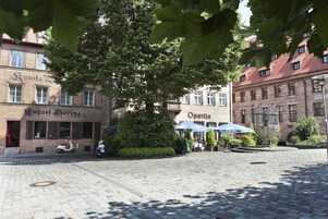 Uma praça de paralelepípedos com uma árvore, um scooter estacionado e edifícios com placas
