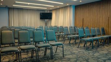 Uma sala de conferências com fileiras de cadeiras, uma grande televisão de tela plana e um tapete estampado.
