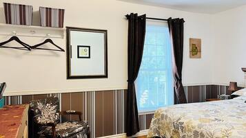 Quarto de hotel com cama, cadeira, escrivaninha, porta-roupas e janela com cortinas.