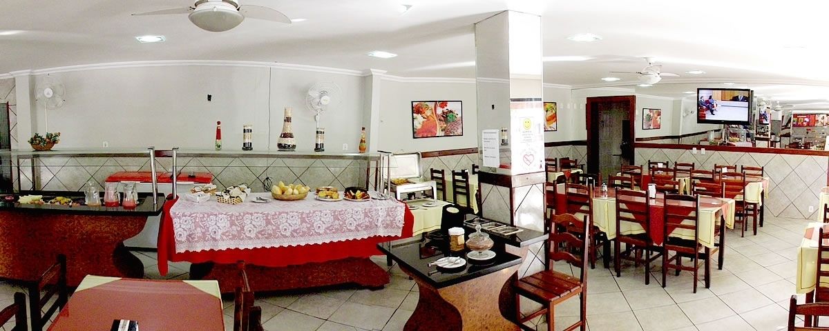 Um restaurante com várias mesas e cadeiras, uma área de buffet com comida e bebidas, e uma tela de televisão.