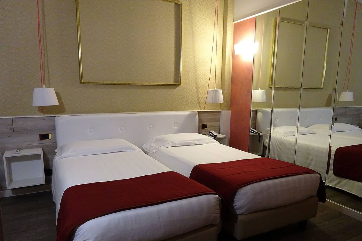Quarto de hotel com duas camas, uma mesinha de cabeceira com abajur, um espelho e uma parede com detalhes em vermelho.