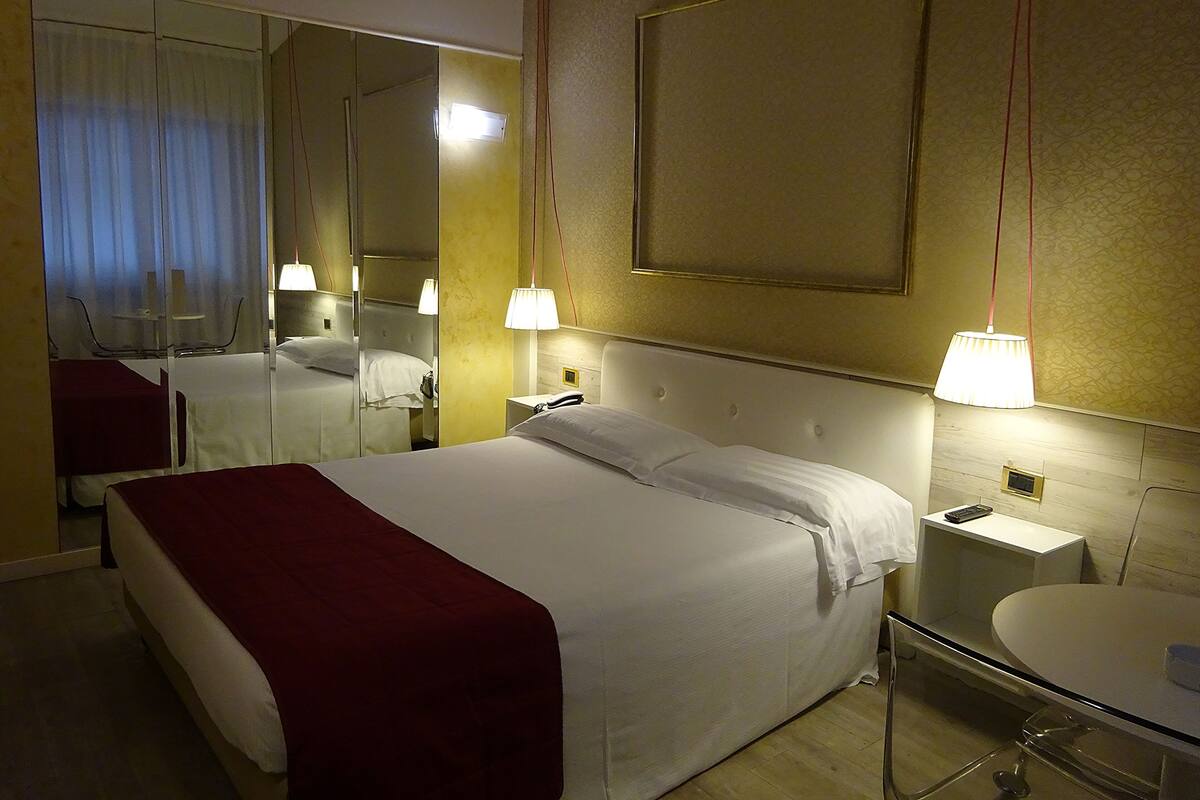 Quarto de hotel com cama grande, luminárias de cabeceira, espelho, mesa de cabeceira com telefone e mesa redonda com cadeira.